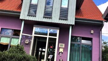 Um hotel chamado "Hotel Ambiente" com telhado de telhas vermelhas, fachada roxa e entrada com piso de tijolos.