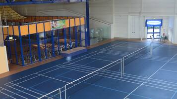 Uma quadra de tênis coberta com piso azul e linhas divisórias brancas, cercada por teto alto com vigas de metal e um revestimento de vidro em um dos lados.