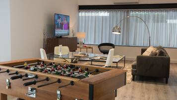 Uma sala de estar com mesa de futebol, televisão, sofá e janela com cortinas.