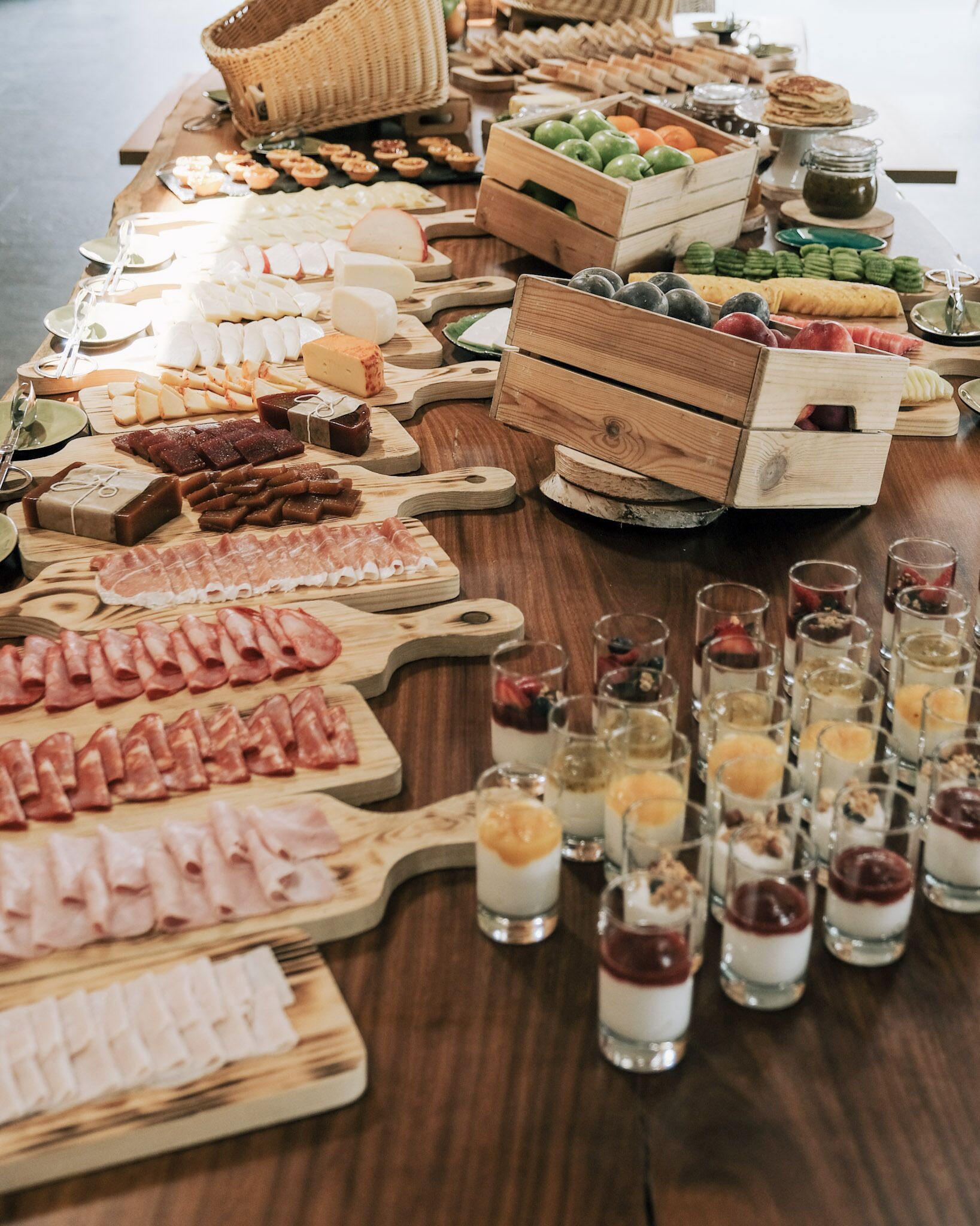Uma longa mesa de madeira repleta de diversos alimentos e bebidas, incluindo queijos, carnes, frutas e pequenas taças.