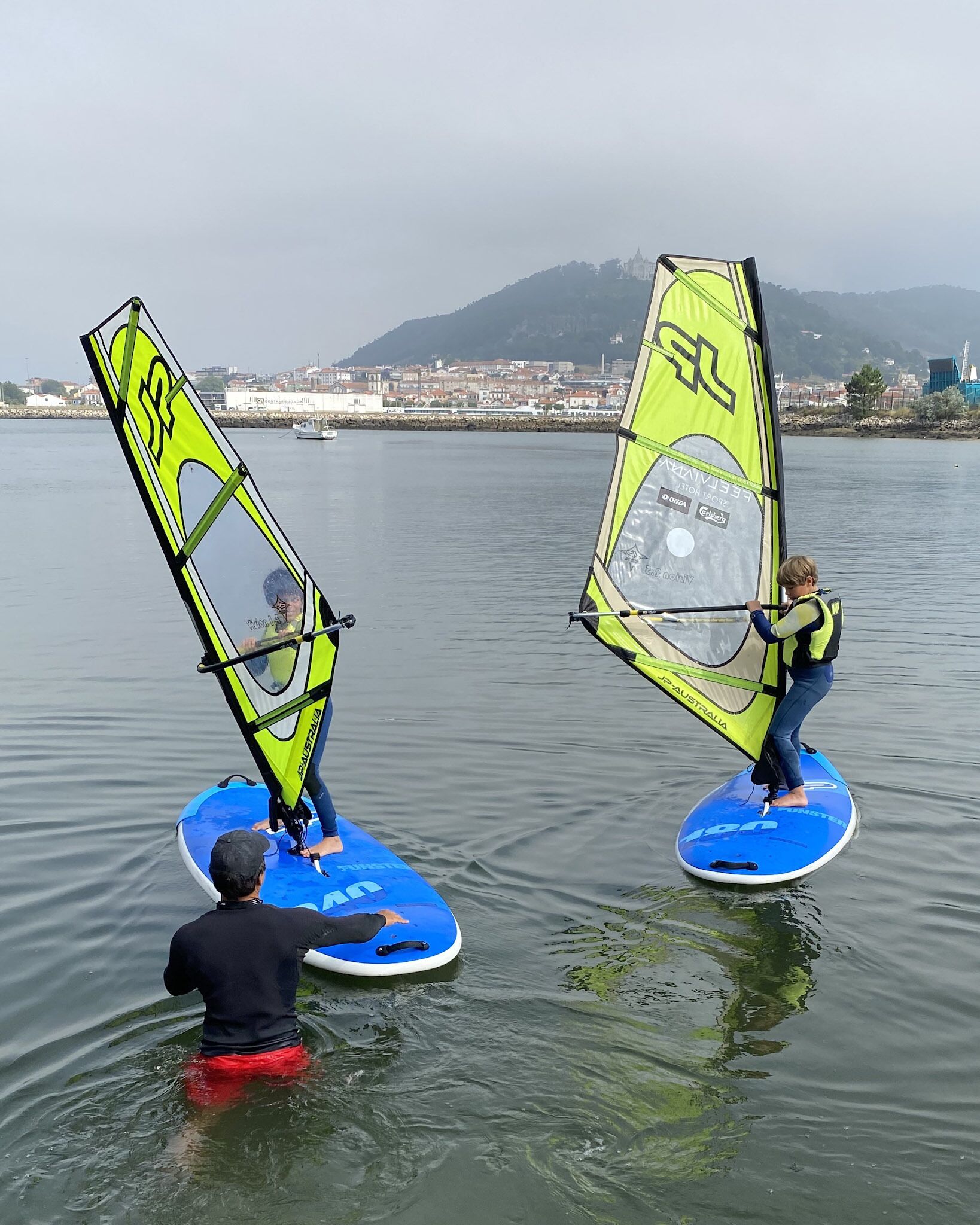 Duas pessoas em um corpo d'água com equipamento de windsurf, uma paisagem urbana ao longe e colinas ao fundo.
