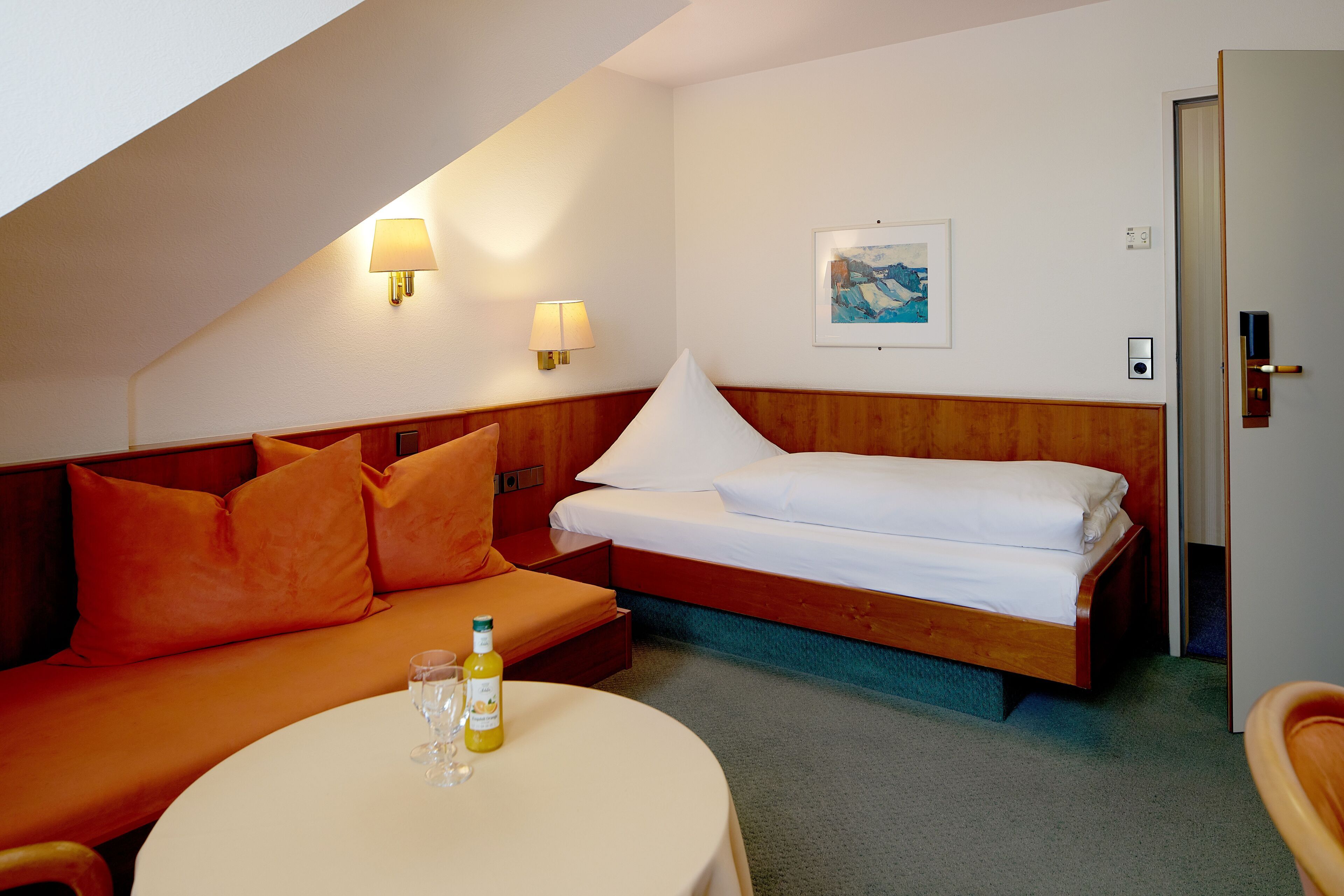 Quarto de hotel com uma cama, um sofá, uma mesa com copos e uma garrafa, e um quadro na parede.