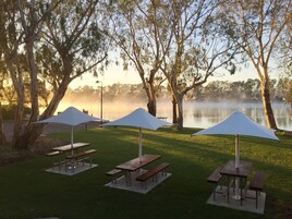Um parque com mesas de piquenique e guarda-sóis brancos, rodeado por árvores e um lago.