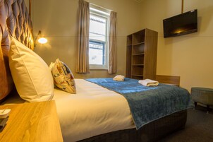 Quarto de hotel com uma cama, uma mesinha de cabeceira de madeira, uma televisão e uma janela com cortinas.