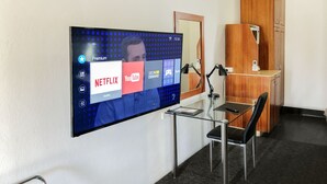 Quarto de hotel com televisão de tela plana na parede, uma escrivaninha com cadeira, um armário de madeira e uma mesinha.