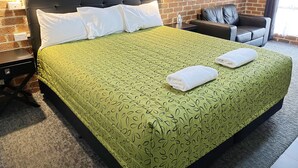 Quarto de hotel com parede de tijolos, cama com colcha verde estampada, duas almofadas brancas, cabeceira preta e uma cadeira de couro preta, além de uma pequena mesa com abajur.