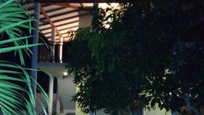 Um edifício de dois andares com um alpendre coberto, um carro estacionado na frente e uma grande árvore ao fundo.