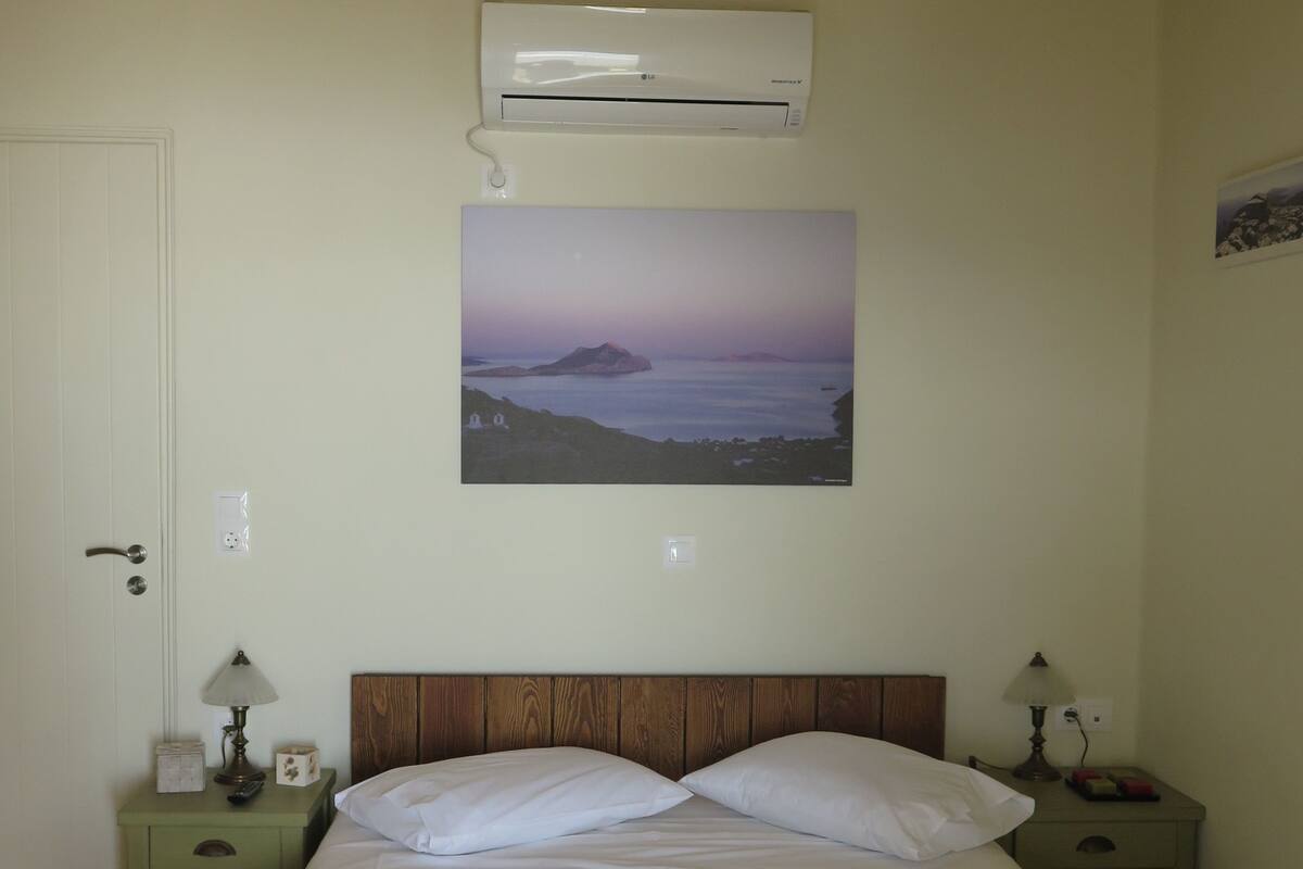Quarto com cama, duas mesas de cabeceira, ventoinha no teto e ar condicionado, além de um quadro na parede.