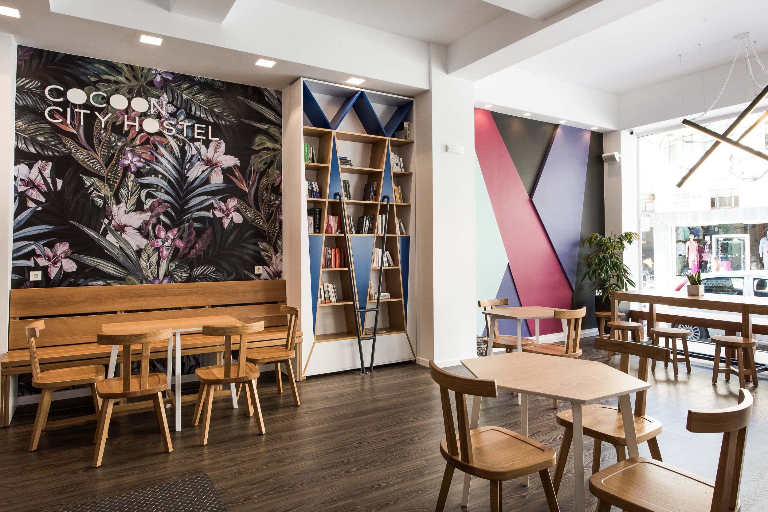 Um espaço interno aconchegante com bancos e mesas de madeira, uma estante de livros e um mural floral vibrante na parede.