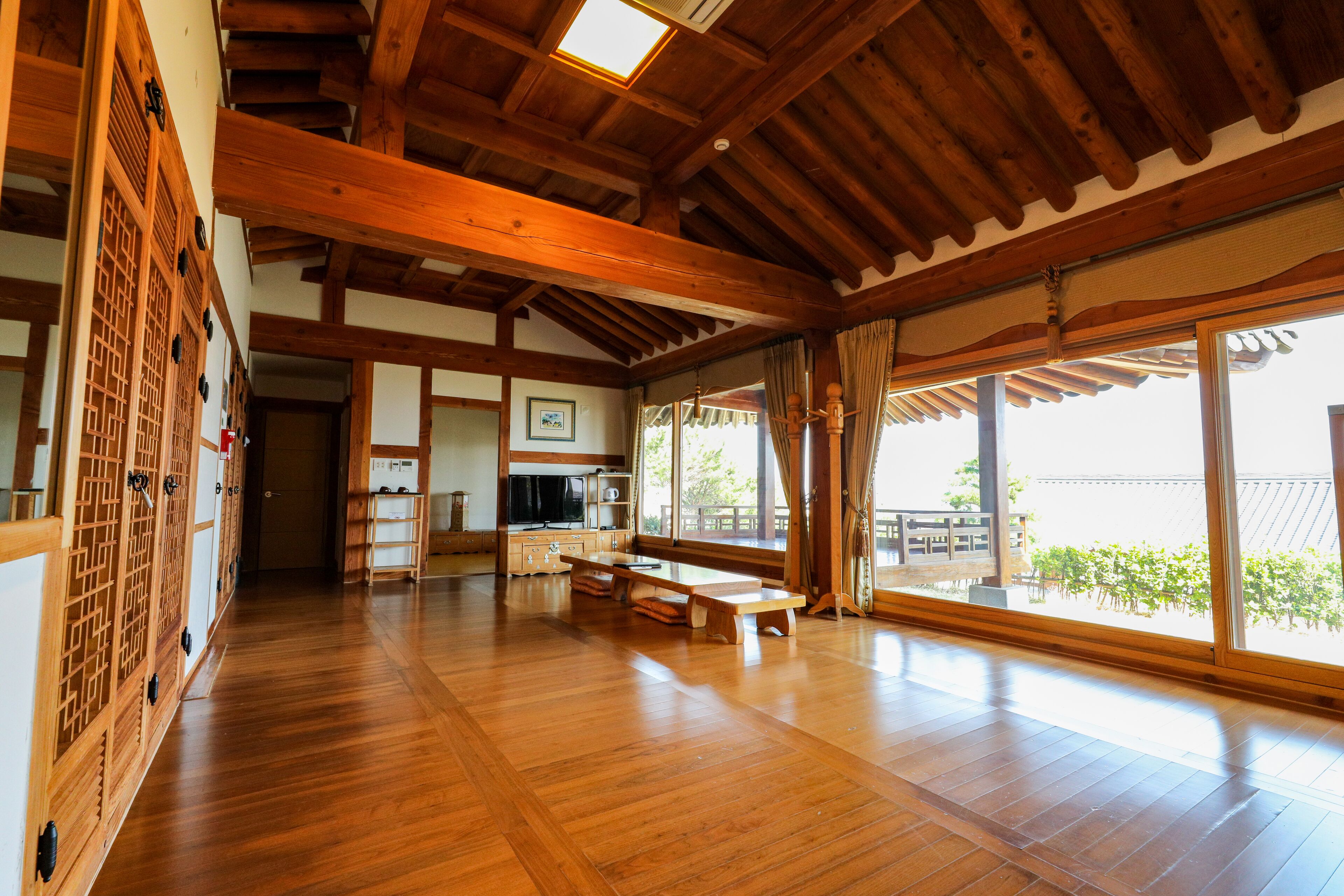 Um quarto amplo com pisos e teto de madeira, uma área de refeições com mesa e bancos, uma cozinha com fogão e armários, e grandes janelas com cortinas.