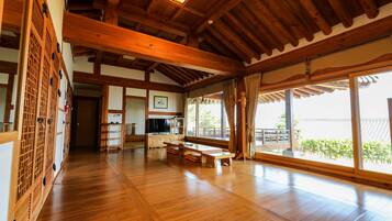 Um quarto amplo com pisos e teto de madeira, uma área de refeições com mesa e bancos, uma cozinha com fogão e armários, e grandes janelas com cortinas.