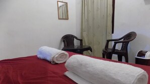 Quarto de hotel com colcha vermelha, duas toalhas brancas enroladas, uma mesinha e duas cadeiras pretas.
