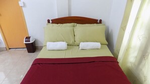 Uma cama de solteiro com cabeceira de madeira, almofadas verdes e brancas e uma colcha vermelha. Há um cesto de lixo à esquerda e uma cortina à direita.