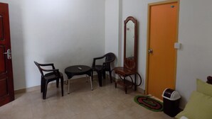 Um quarto com duas cadeiras de madeira, uma mesa pequena, um espelho, uma porta e um cesto de lixo.
