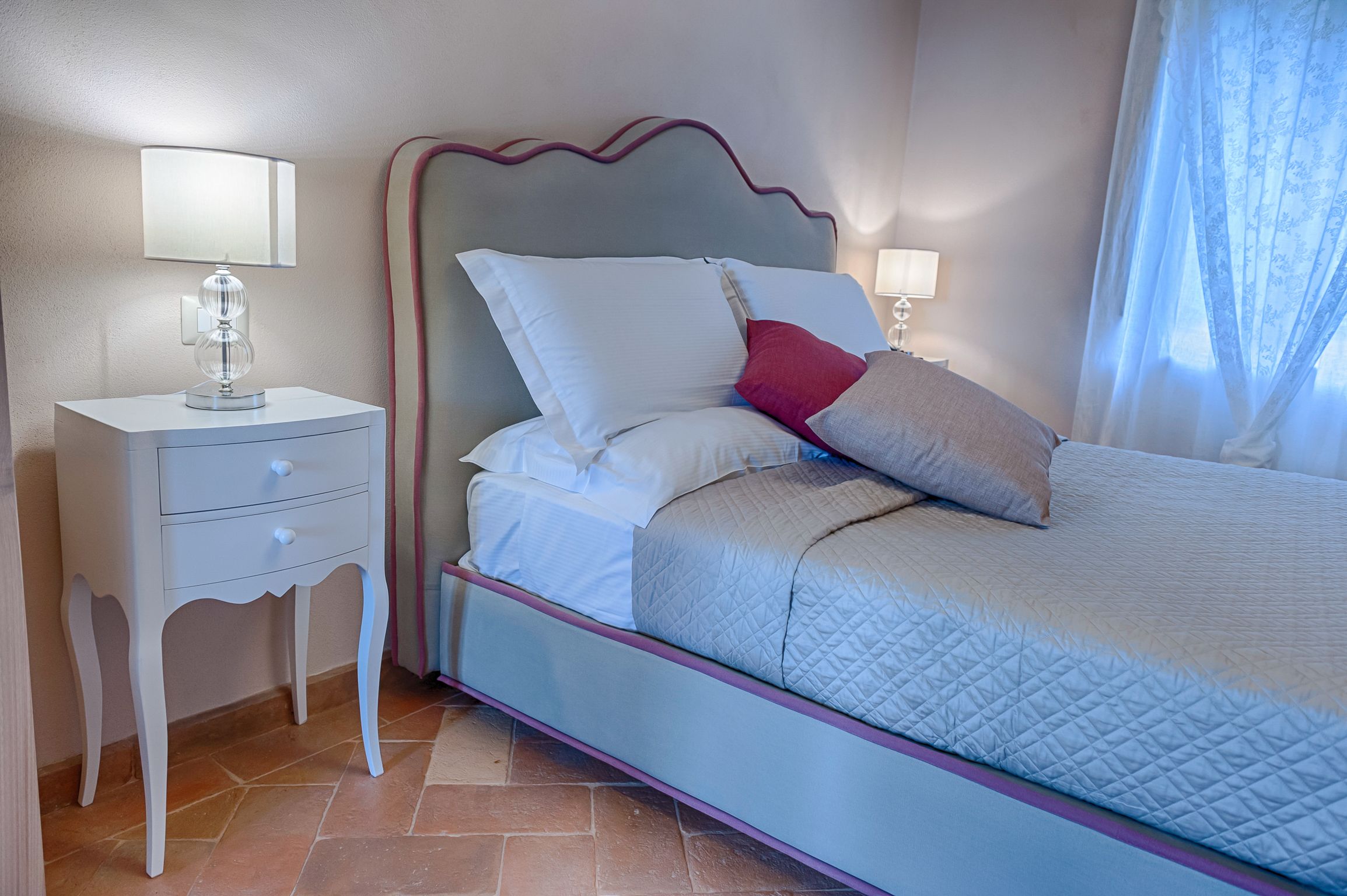 Uma cama bem arrumada com um cobre acolchoado, uma mesa de cabeceira com abajur e um criado-mudo com gavetas.
