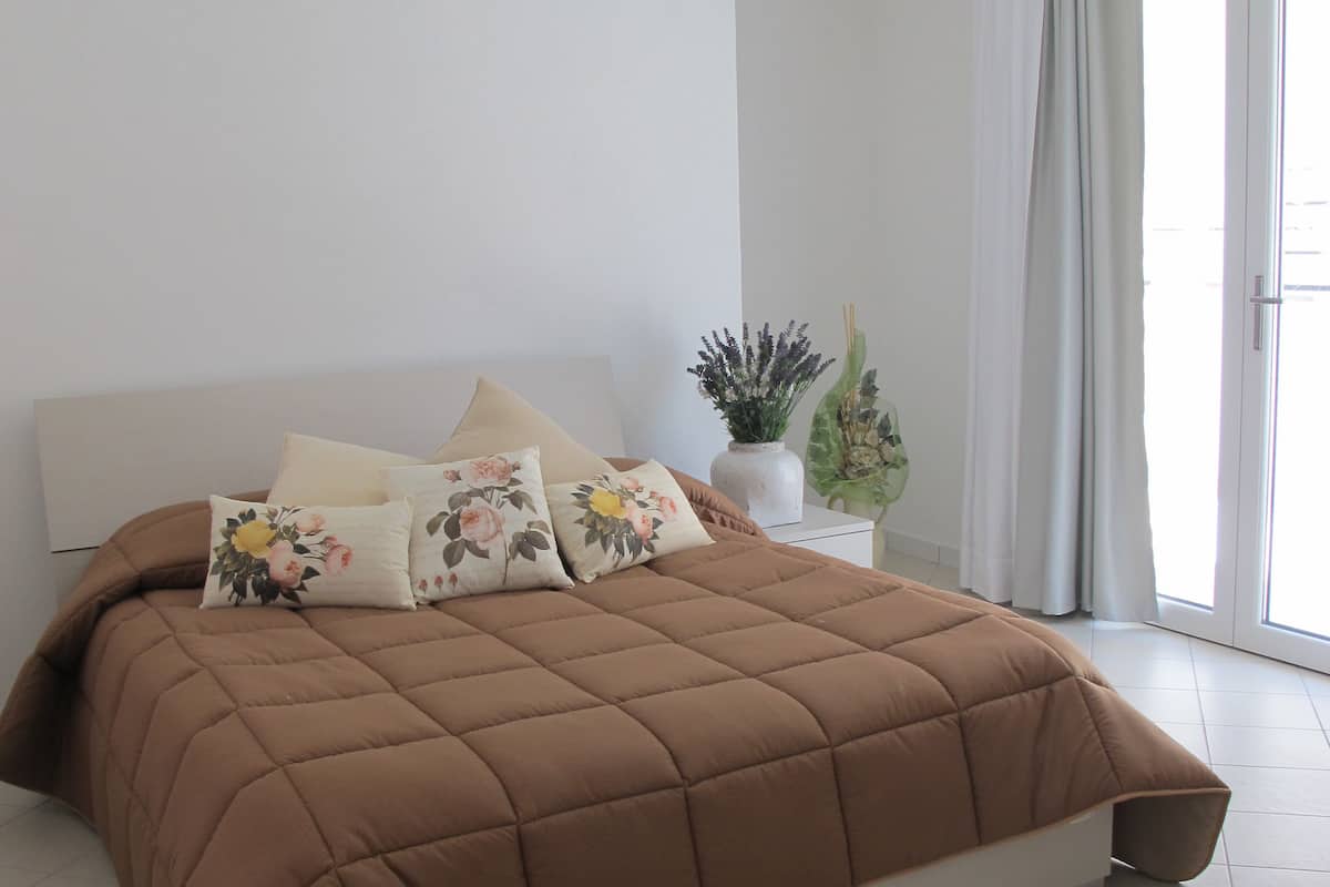 Quarto com cama grande, mesa de cabeceira, planta em vaso e vista para o exterior através de uma janela com cortinas.