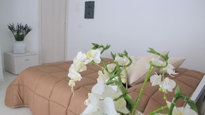Quarto com cama, mesa de cabeceira, um vaso com flores brancas e um painel de interruptores na parede.