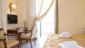 Quarto de hotel com uma cama, duas cadeiras, uma escrivaninha, televisão e janela com cortinas.