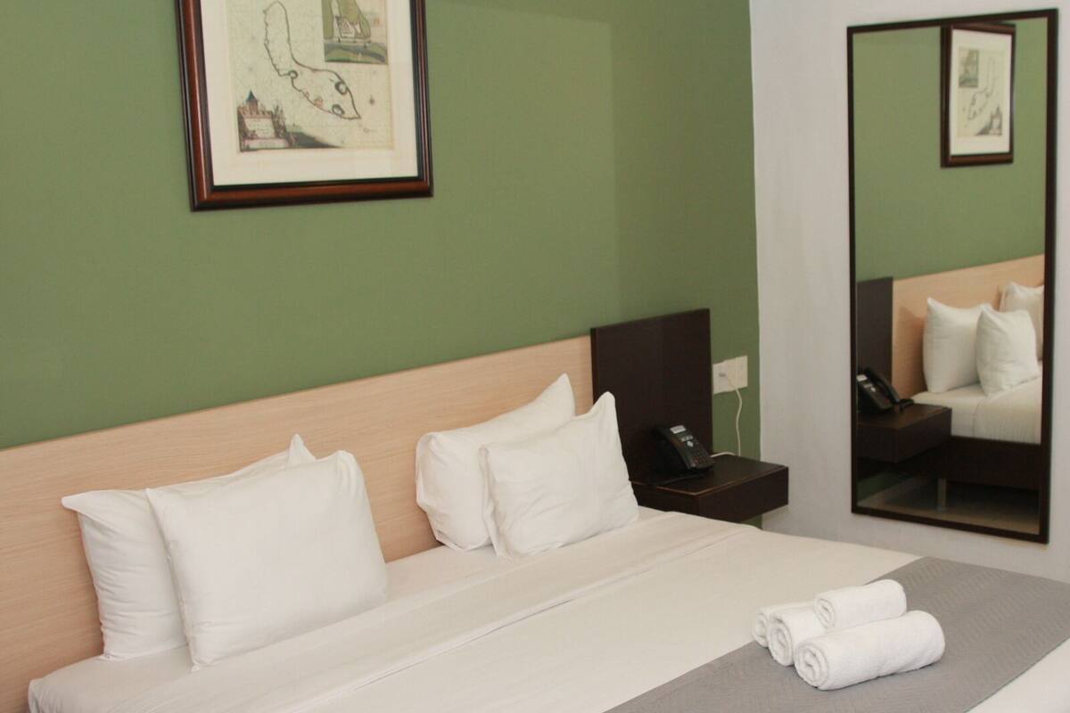 Quarto de hotel com cama, almofadas, mesa de cabeceira com telefone, espelho e quadro na parede.