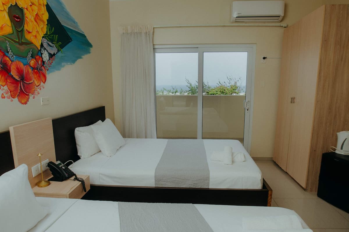 Quarto de hotel com duas camas, um quadro na parede, ar condicionado e vista pela porta de vidro.