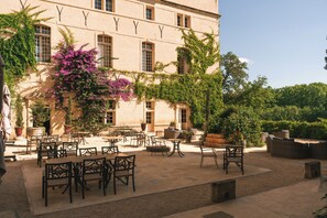 Pátio externo com mesas e cadeiras, rodeado por um edifício com grandes janelas e vegetação.
