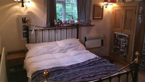 Quarto com cama, janela com cortinas, radiador, escrivaninha com luminária e estante de livros.