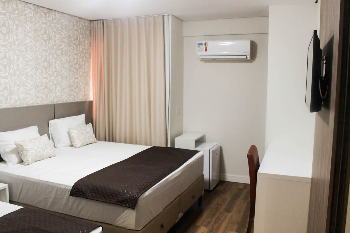 Quarto de hotel com cama, mesa de cabeceira, cadeira, ar condicionado e televisão fixada na parede.