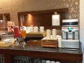 Uma mesa de pequeno-almoço com máquinas de café, uma apresentação de frutas e variedade de chávenas e copos.