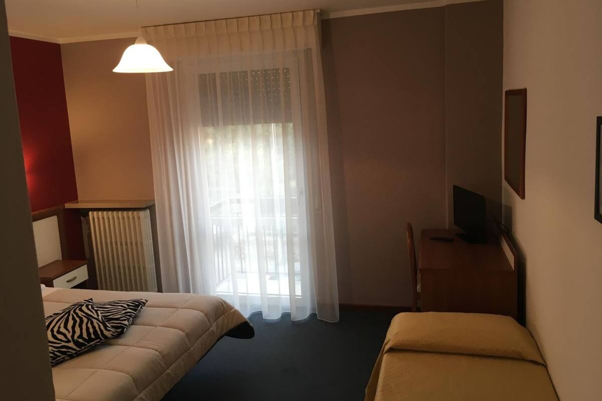 Quarto de hotel com cama, sofá, escrivaninha com computador e janela com cortinas leves.