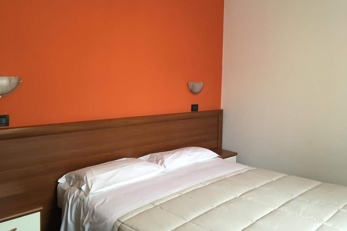 Cama com cabeceira de madeira, roupa de cama branca e parede com detalhe laranja.