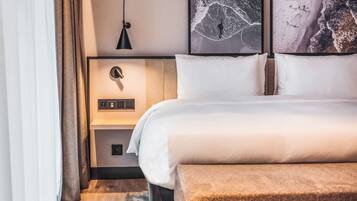 Quarto de hotel moderno com uma cama grande, uma mesinha de cabeceira, duas mesas laterais e dois quadros na parede.
