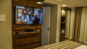 Quarto de hotel com uma cama, televisão presa a um armário de madeira e um guarda-roupa com espelho.