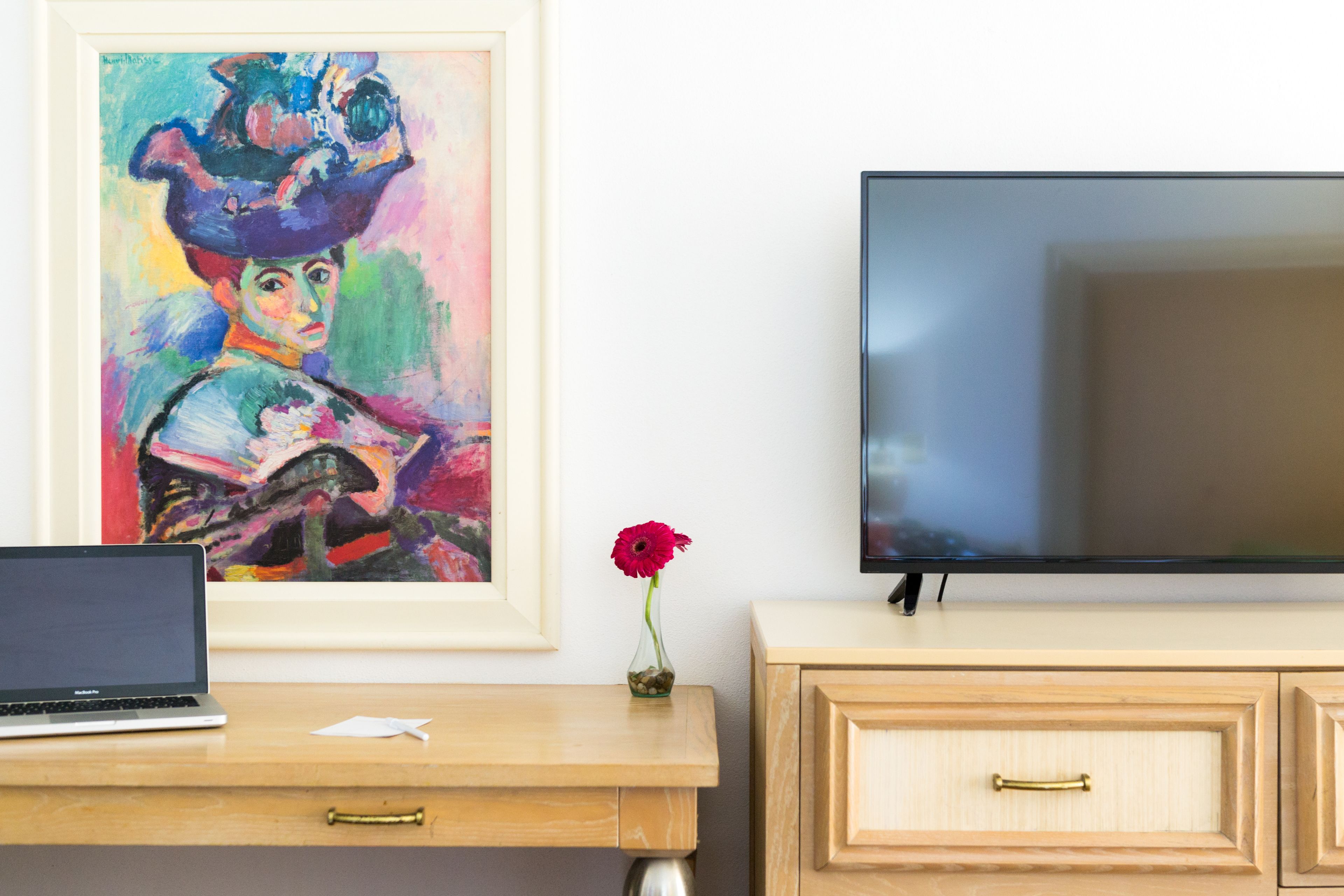 Um quarto com uma escrivaninha de madeira, um laptop, um quadro emoldurado, um vaso com uma flor vermelha e uma televisão em um suporte.