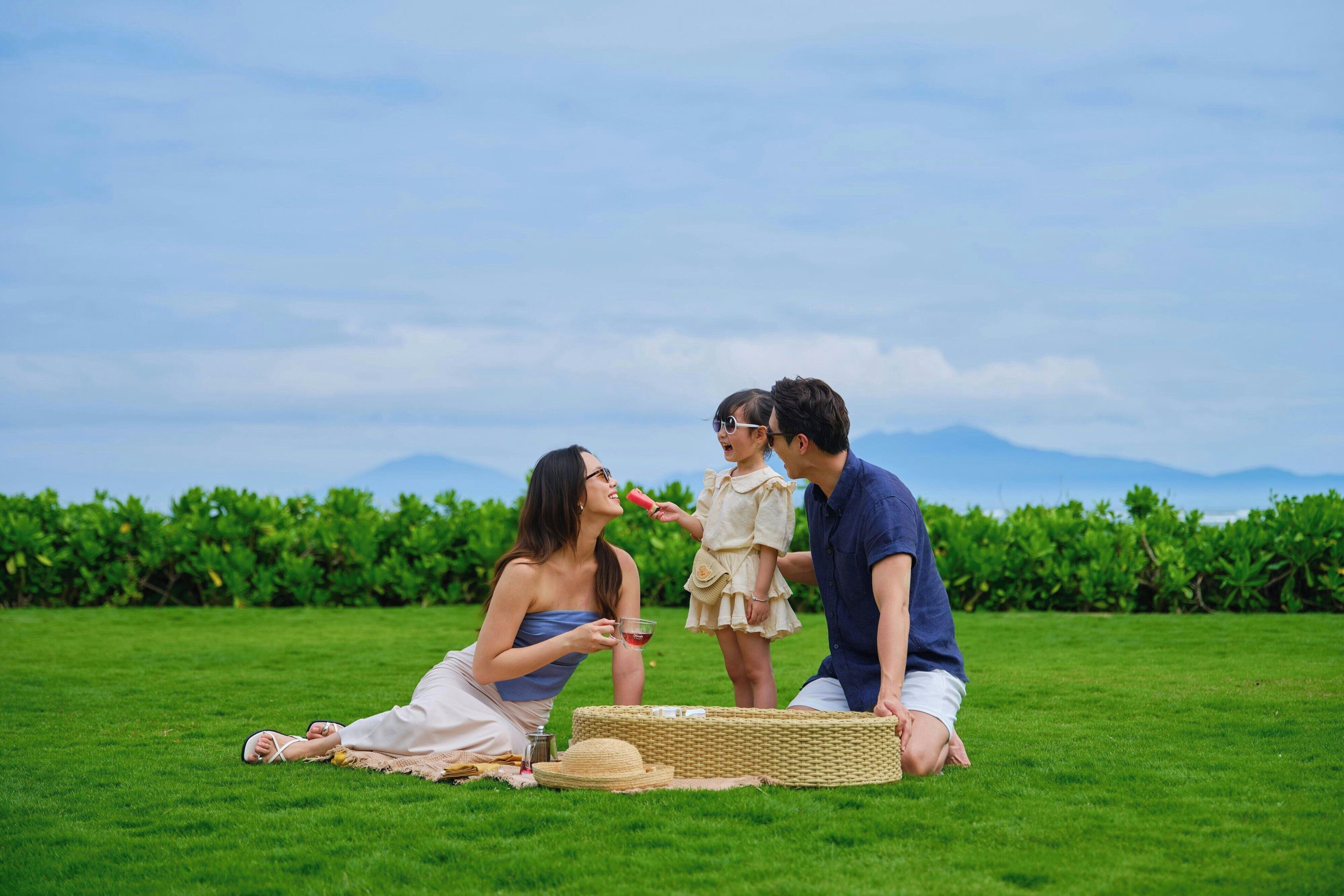 Uma família fazendo piquenique em um campo gramado, com céu claro e montanhas ao longe.