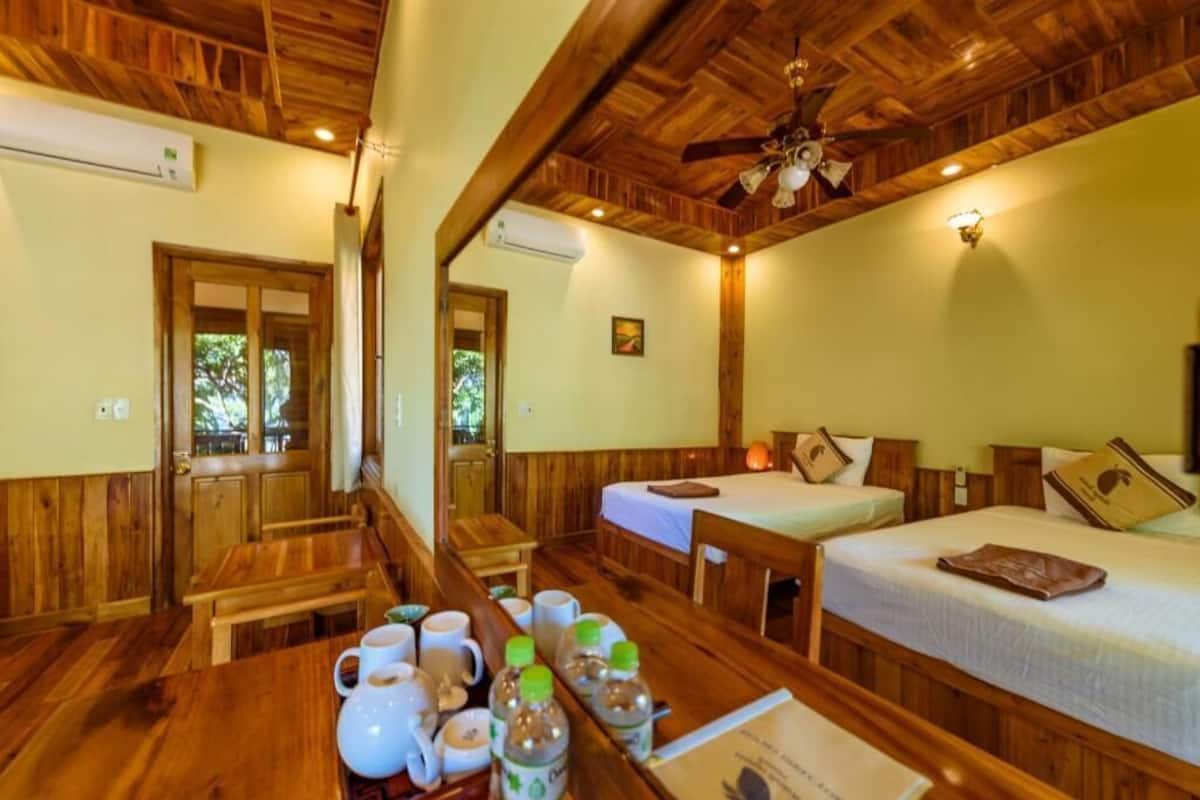 Quarto com piso de madeira, duas camas, uma mesa de jantar com canecas e garrafas, e um ventilador de teto.