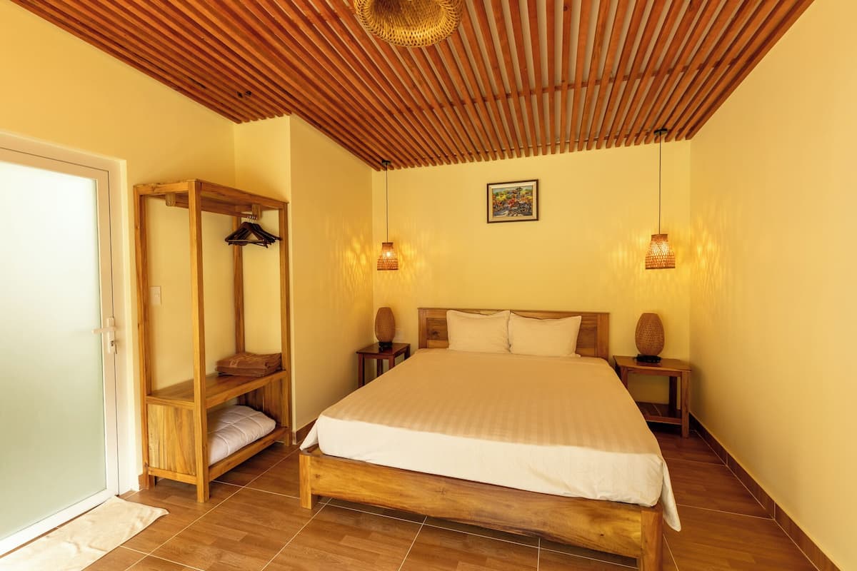 Quarto com cama de madeira, guarda-roupa de madeira, duas mesas de cabeceira com luminárias e uma porta à esquerda.