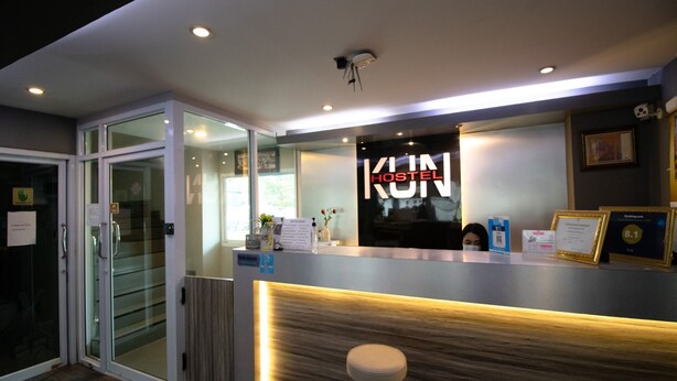 Um balcão de recepção moderno com entrada por porta de vidro, uma parede de destaque em madeira e uma placa com a inscrição "KUN HOSTEL".