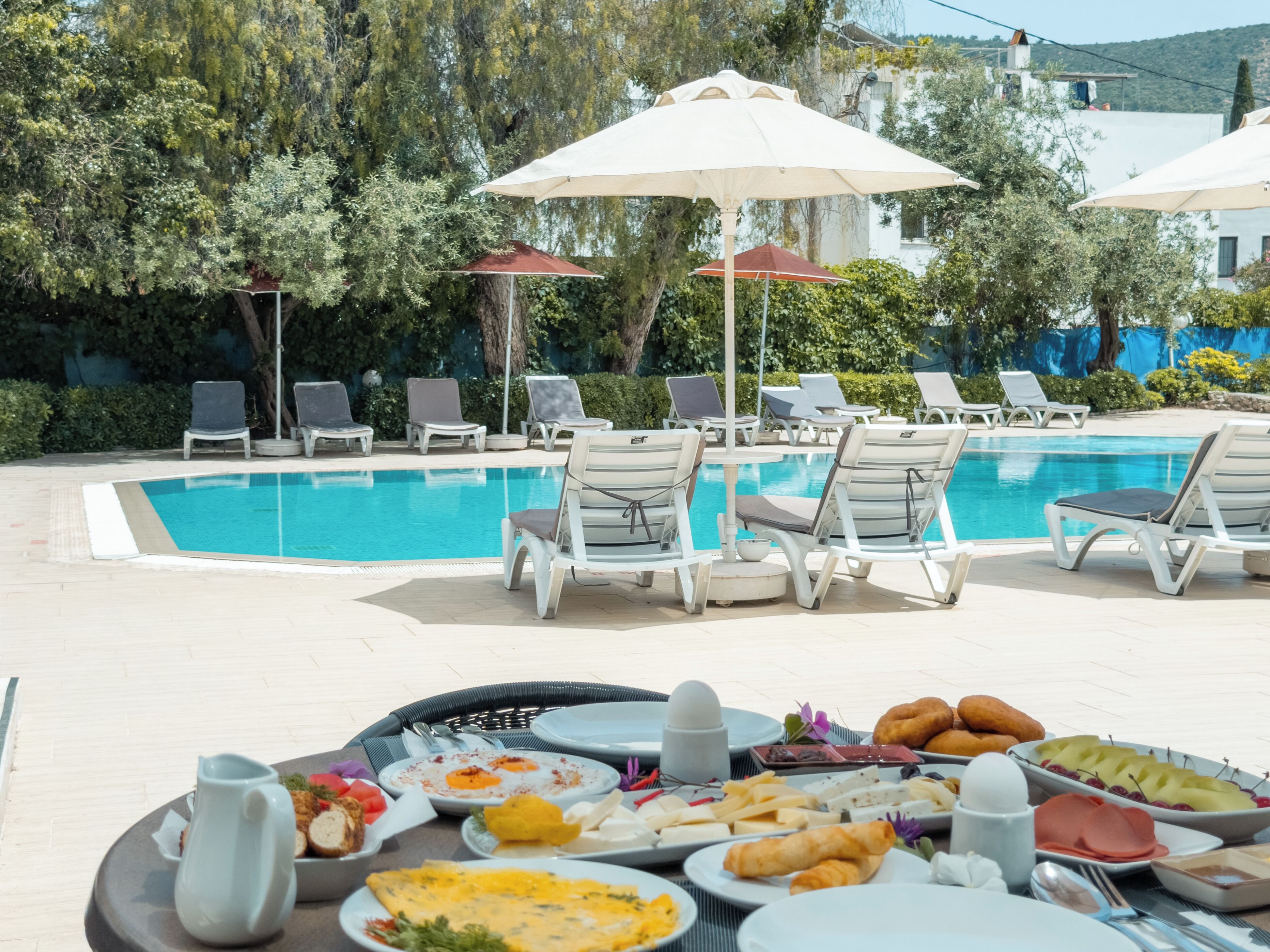 Uma área de refeições à beira da piscina com espreguiçadeiras e guarda-sóis, piscina e mesa com itens de café da manhã.