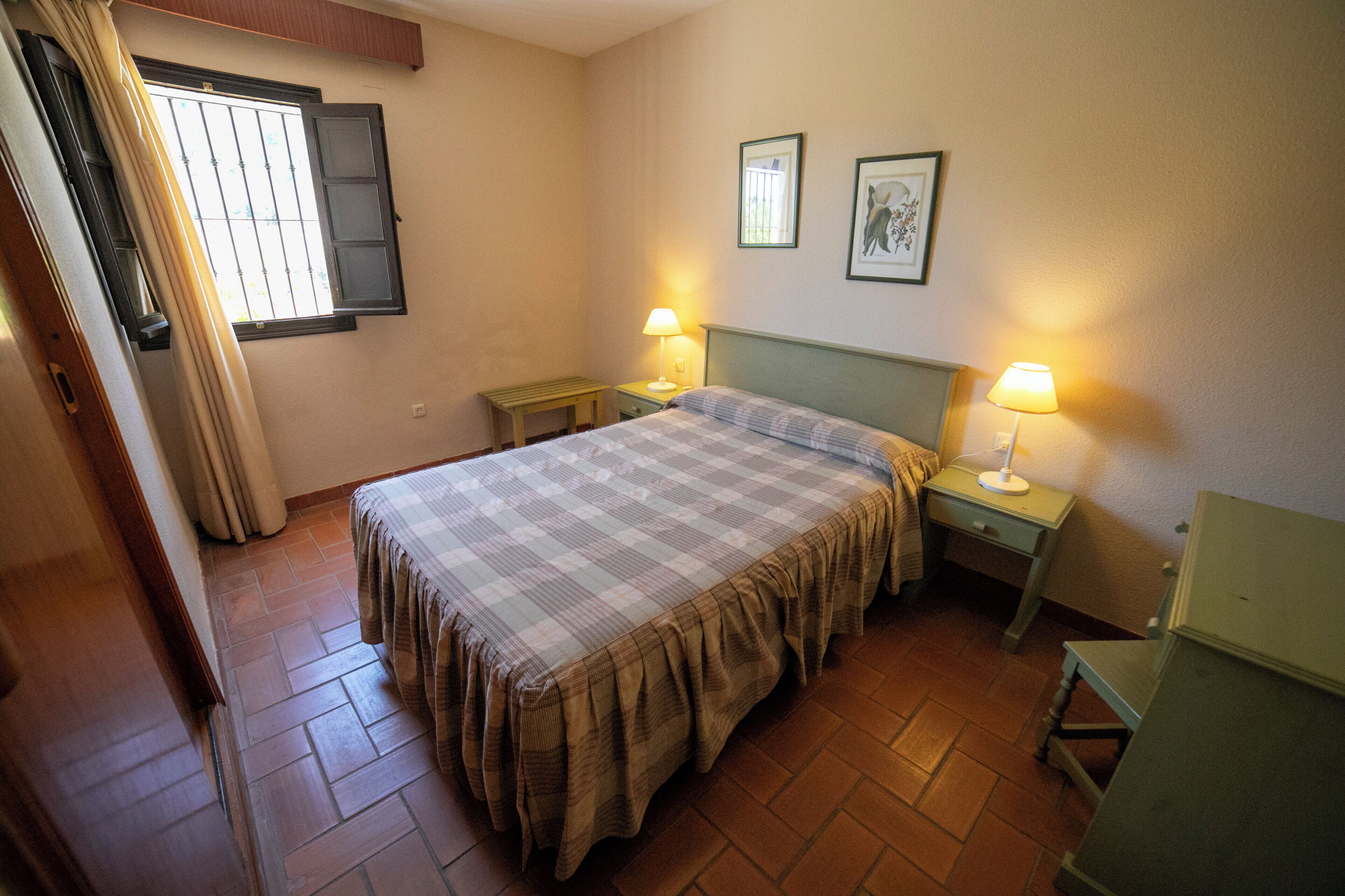 Quarto com uma cama, duas mesas de cabeceira com luminárias, uma janela com cortinas e duas imagens emolduradas na parede.