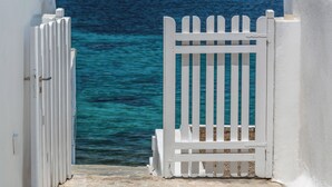 Um portão branco dá acesso a uma área costeira com águas azul-claras e montanhas ao fundo.
