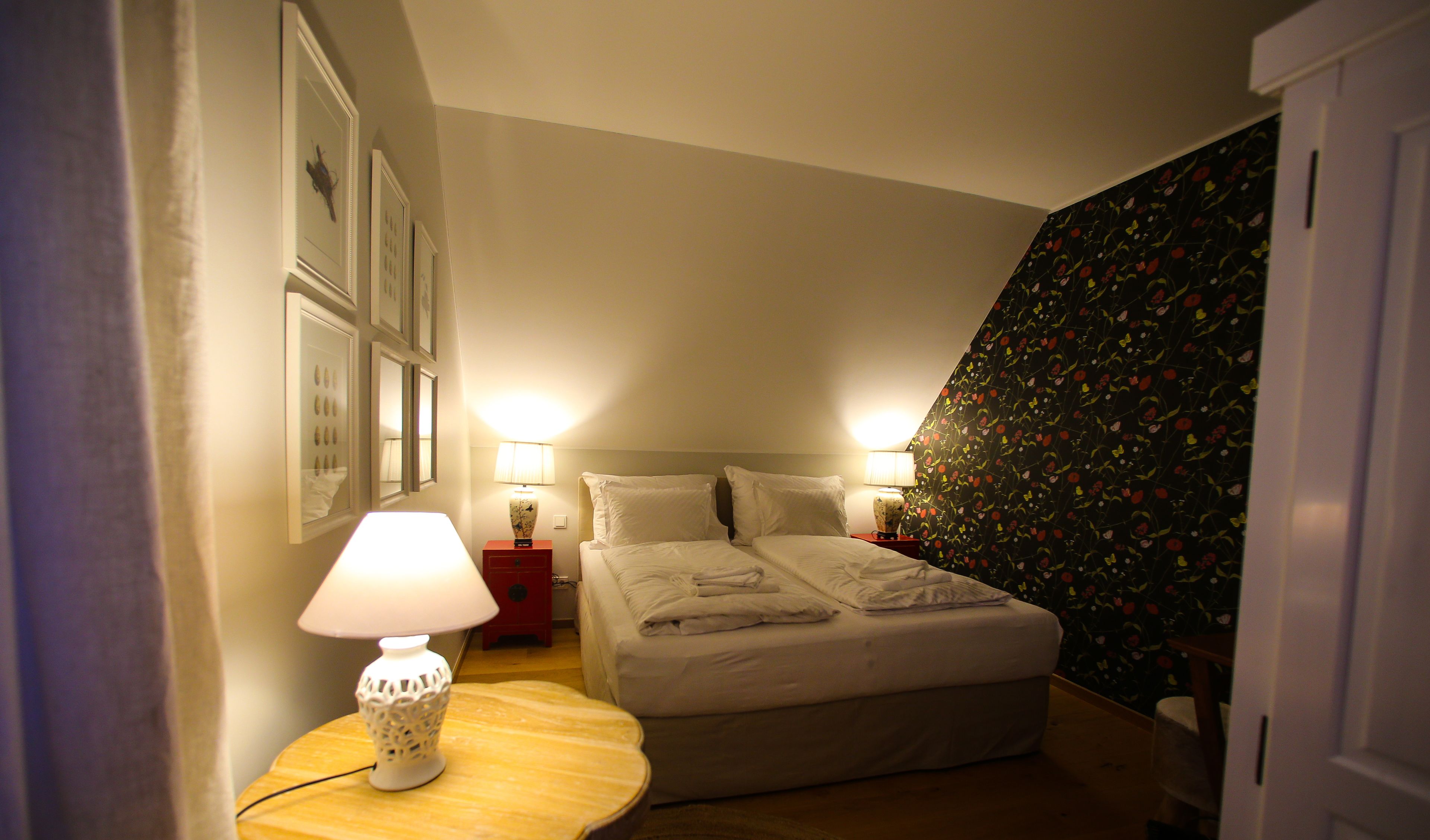 Quarto com cama, duas luminárias de cabeceira, mesa de cabeceira e parede com papel de parede floral.