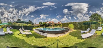 Uma piscina circular com espreguiçadeiras e guarda-sóis, cercada por um gramado bem cuidado e sebes.