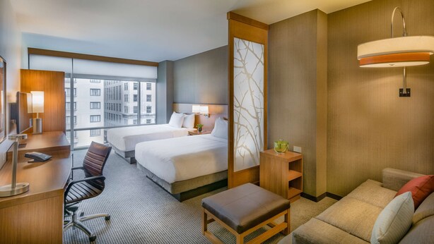 Quarto de hotel com duas camas, uma escrivaninha com telefone, uma cadeira, um sofá, uma pequena mesa com uma planta e uma janela grande com vista para a cidade.