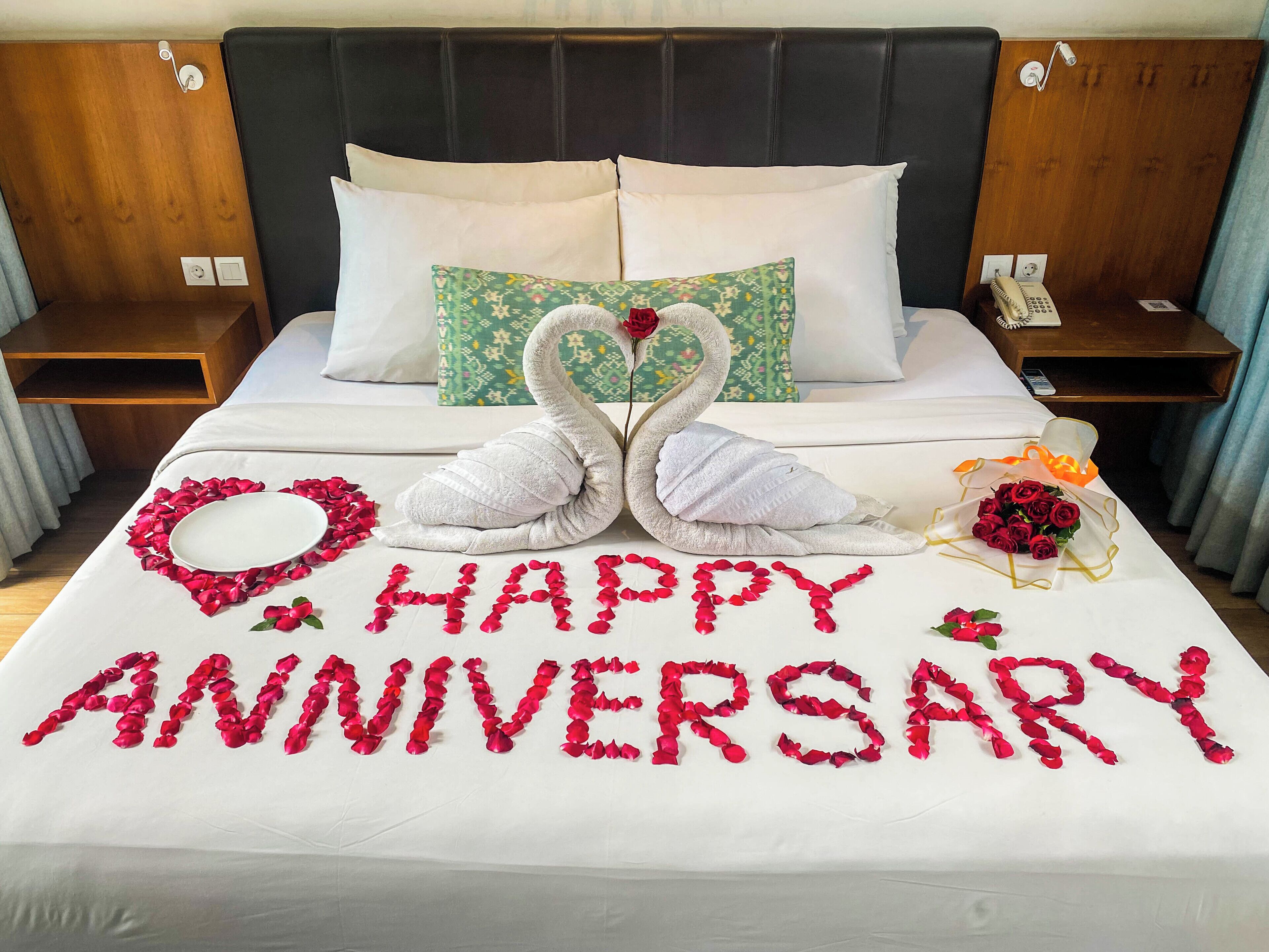 Uma cama com arranjo de toalha em formato de cisne e a frase 'HAPPY ANNIVERSARY' escrita com pétalas de rosa.