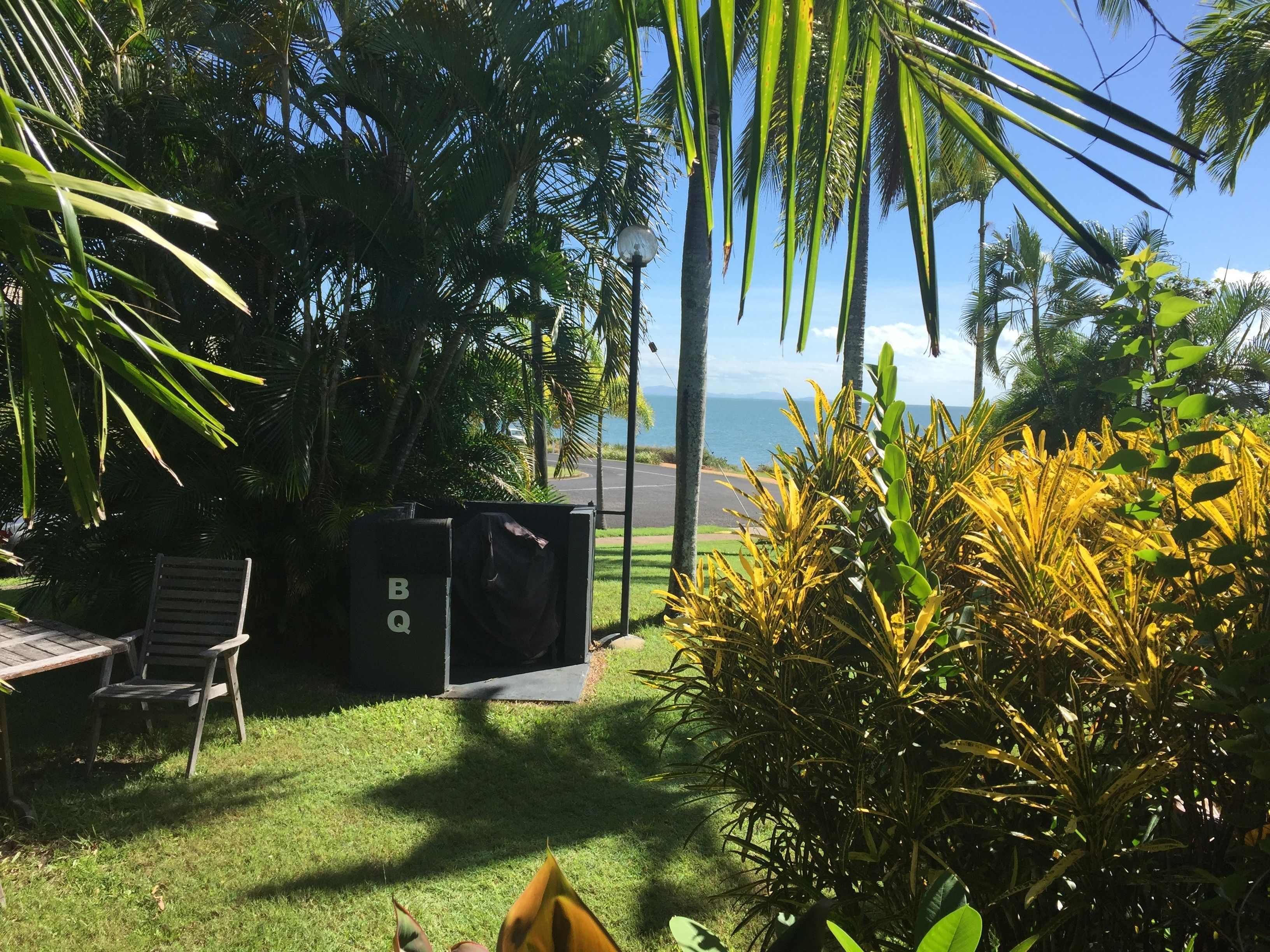 Um jardim com um banco de madeira, uma estrutura preta identificada como 'BQ', e vista para o oceano.