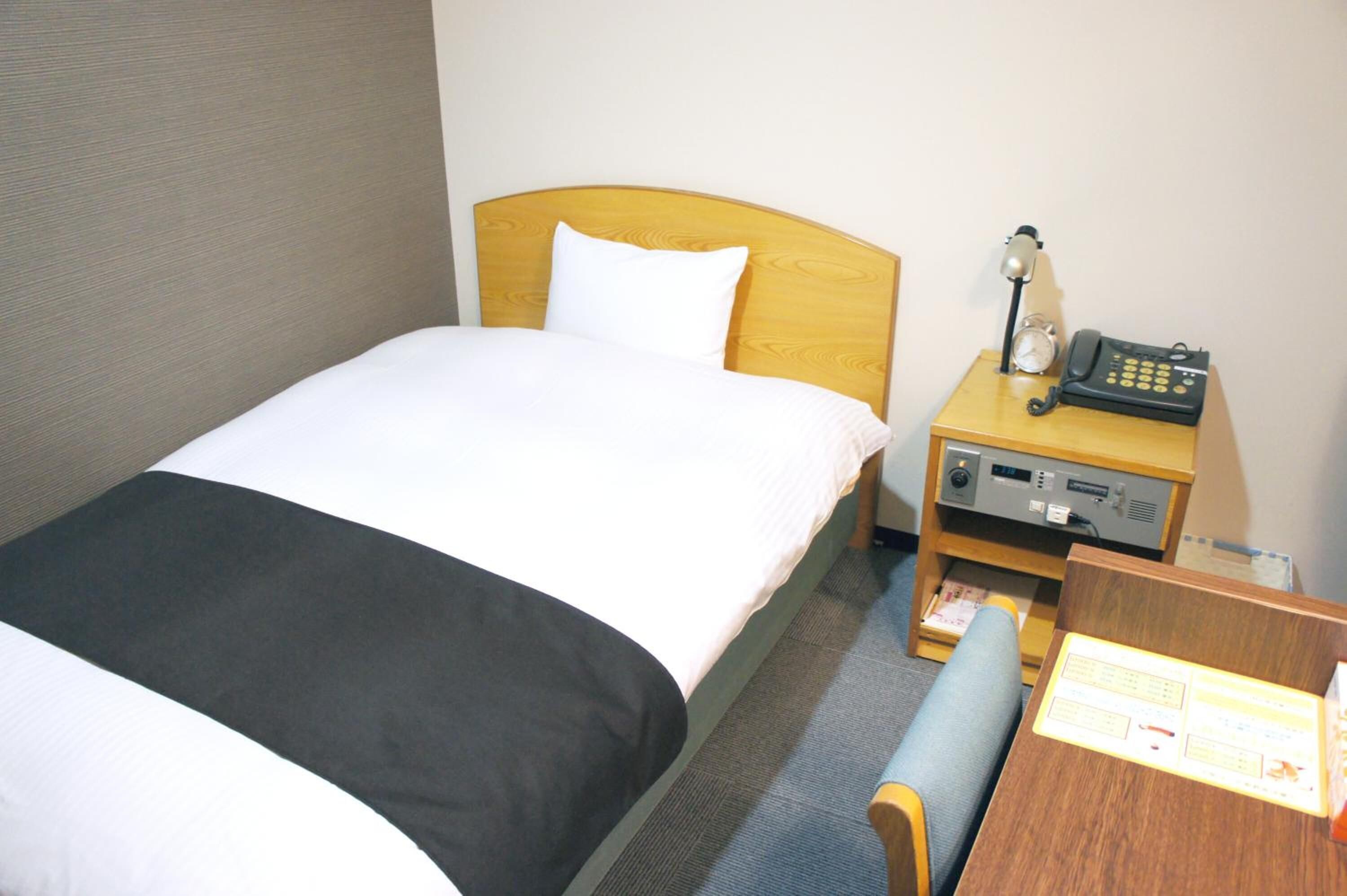 Quarto de hotel com uma cama de solteiro, mesa de cabeceira com telefone, abajur e pequena escrivaninha com cadeira.