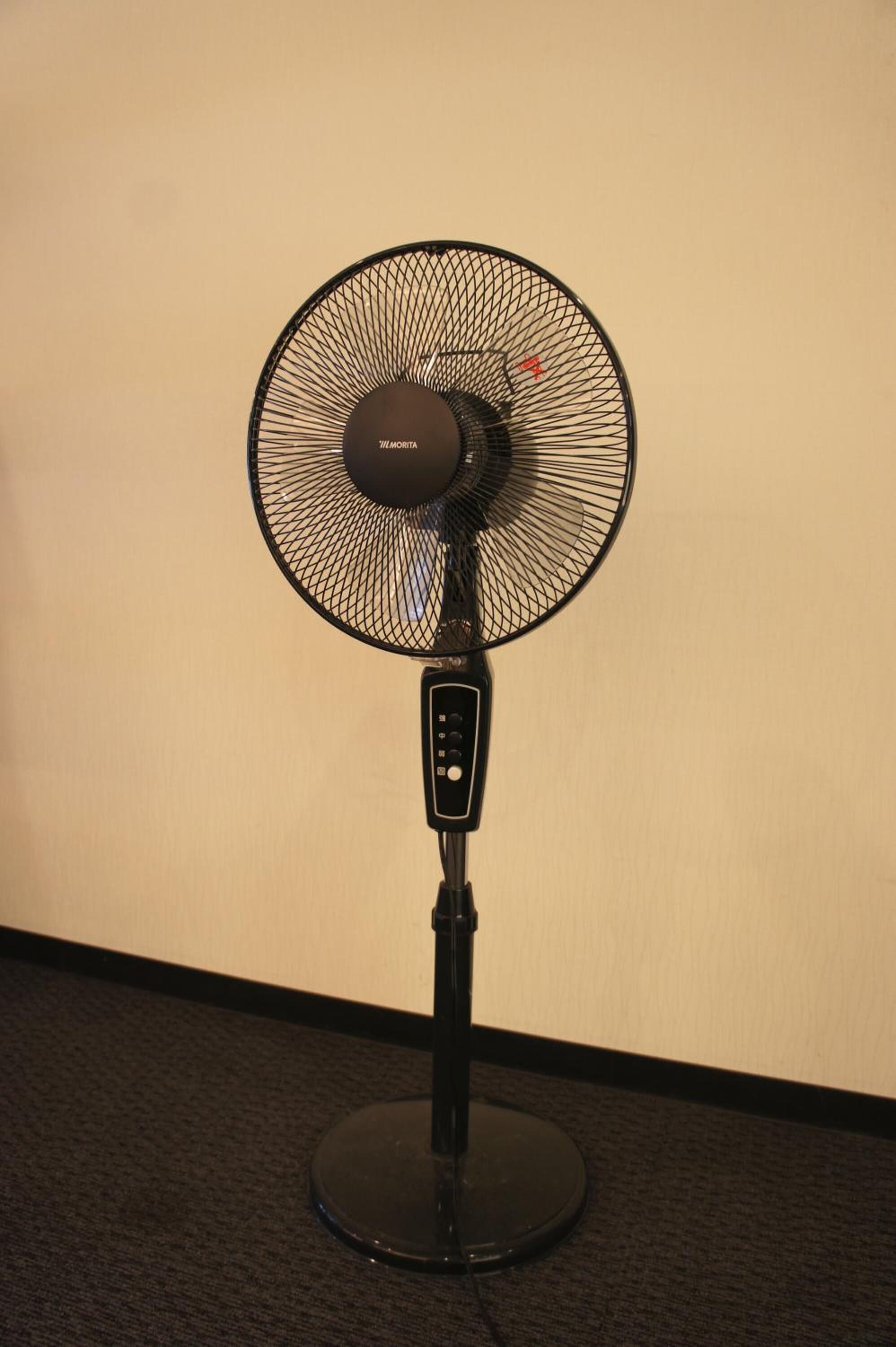 Um ventilador elétrico de coluna, com painel de controle, posicionado sobre um piso coberto de carpete, em frente a uma parede lisa.