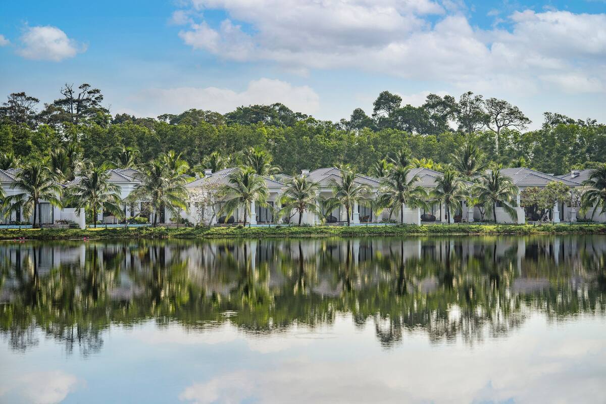 Fileira de edifícios brancos com telhados cinzentos, rodeada por palmeiras, refletindo em águas calmas.