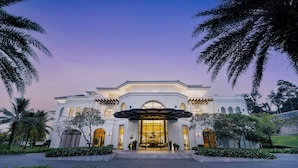 Um edifício grande e elegante com uma entrada imponente, rodeado por vegetação exuberante e palmeiras.
