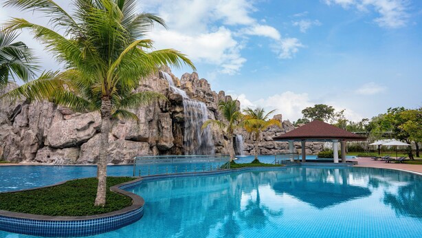 Piscina com cascata, cercada por palmeiras e um gazebo.